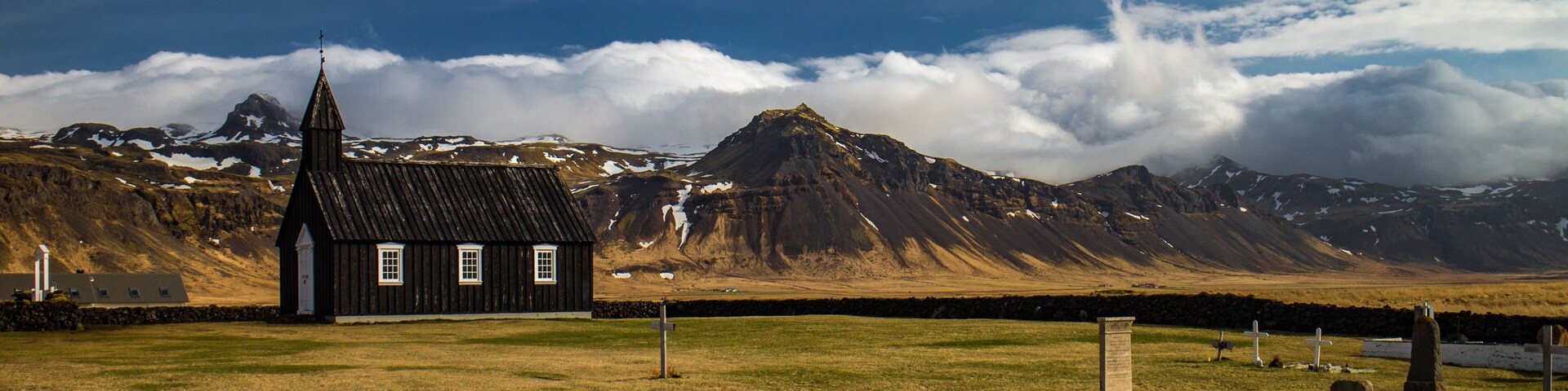 Uma igreja negra com um campanário, uma cruz de pedra e um cemitério com lápides em um campo gramado, com montanhas ao fundo.