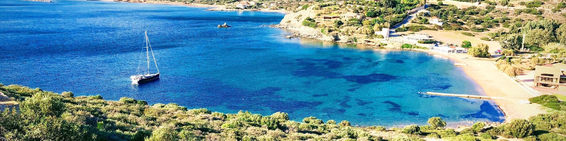 Paisagem costeira com mar azul límpido, um veleiro, terreno rochoso e edifícios espalhados.