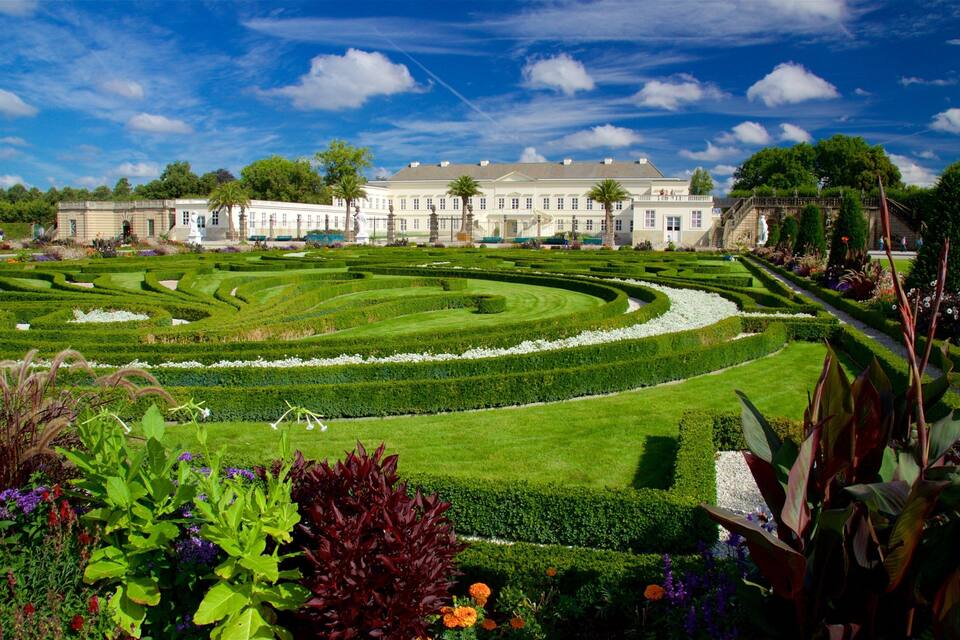 Um jardim formal com sebes geometricamente dispostas, um edifício branco imponente e estátuas.