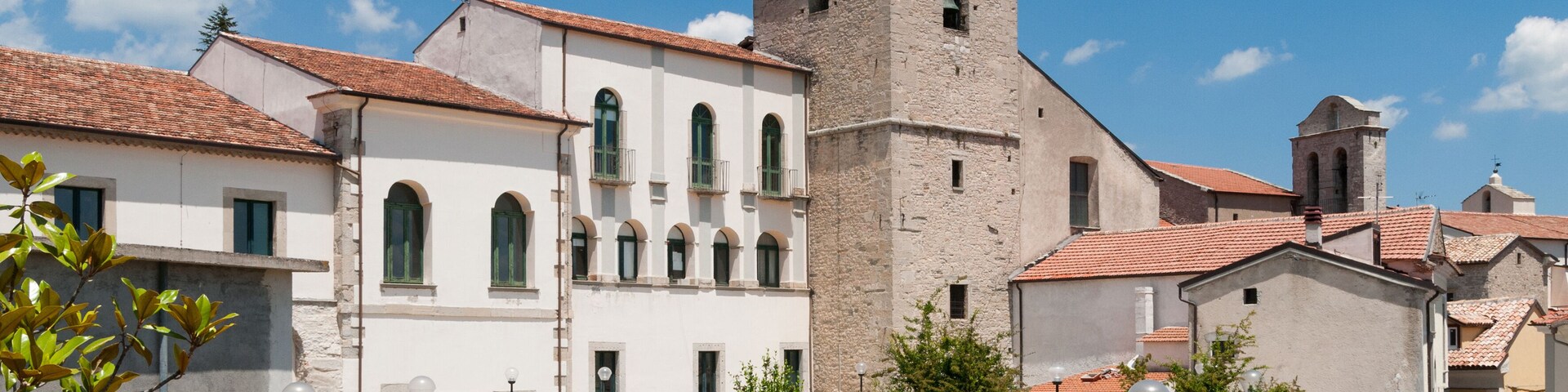 Um edifício histórico com uma torre do relógio, rodeado por um pequeno pátio com árvores e um caminho pavimentado.