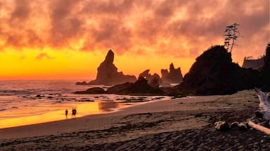 Praia com formações rochosas silhuetadas contra o céu do pôr do sol.