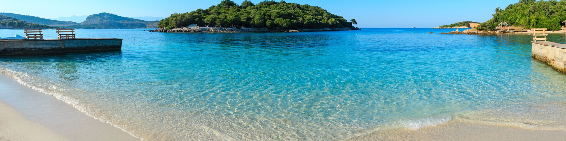 Mar azul transparente com uma praia de areia, um cais de madeira e uma pequena ilha com árvores.