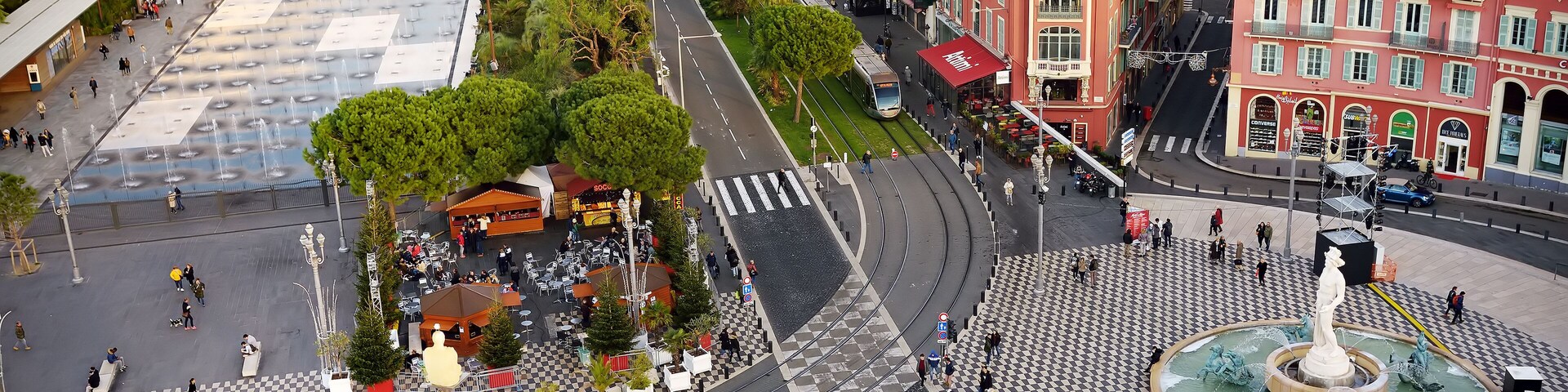 Uma praça urbana com uma fonte, trilhos de bonde e edifícios ao redor.