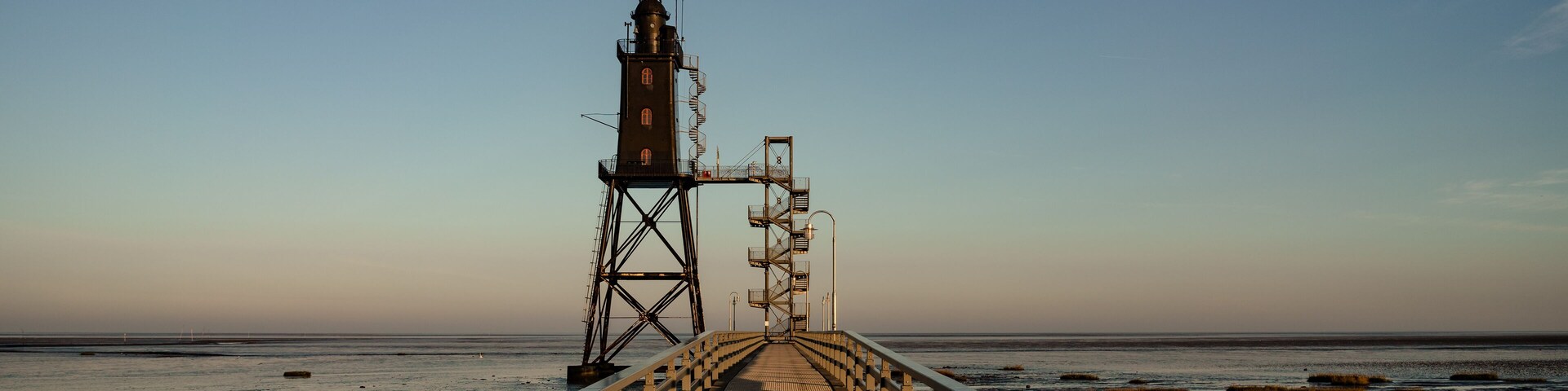 Um cais que leva a um farol com uma bandeira, céu claro e uma praia com zona úmida.