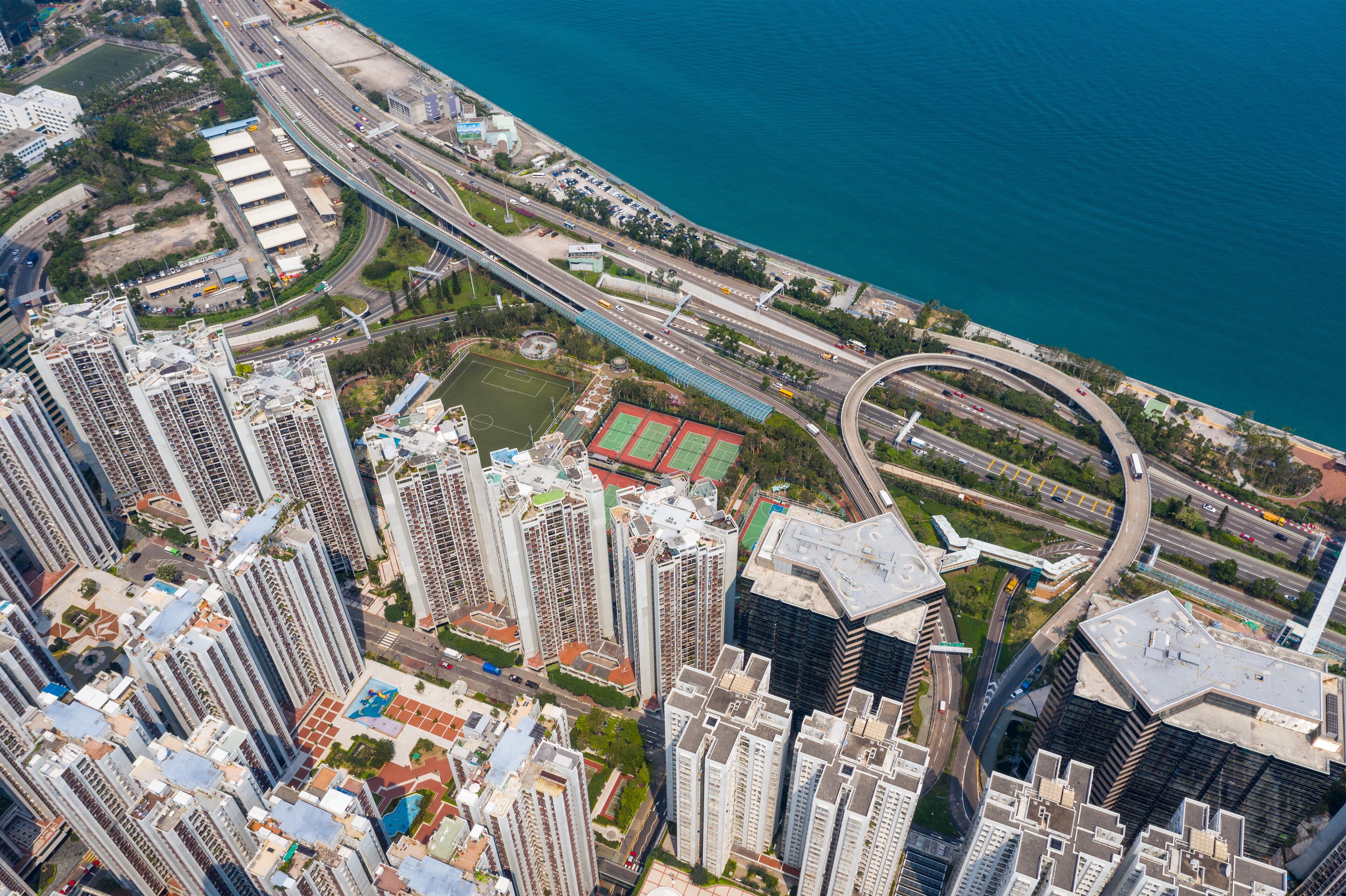 Uma área urbana com edifícios altos, uma estrada sinuosa, um campo esportivo e um corpo d'água.
