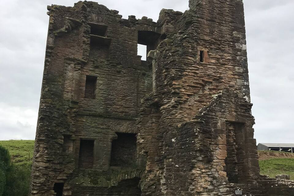 Ruínas de um castelo de pedra, com várias janelas e um campo gramado em primeiro plano.