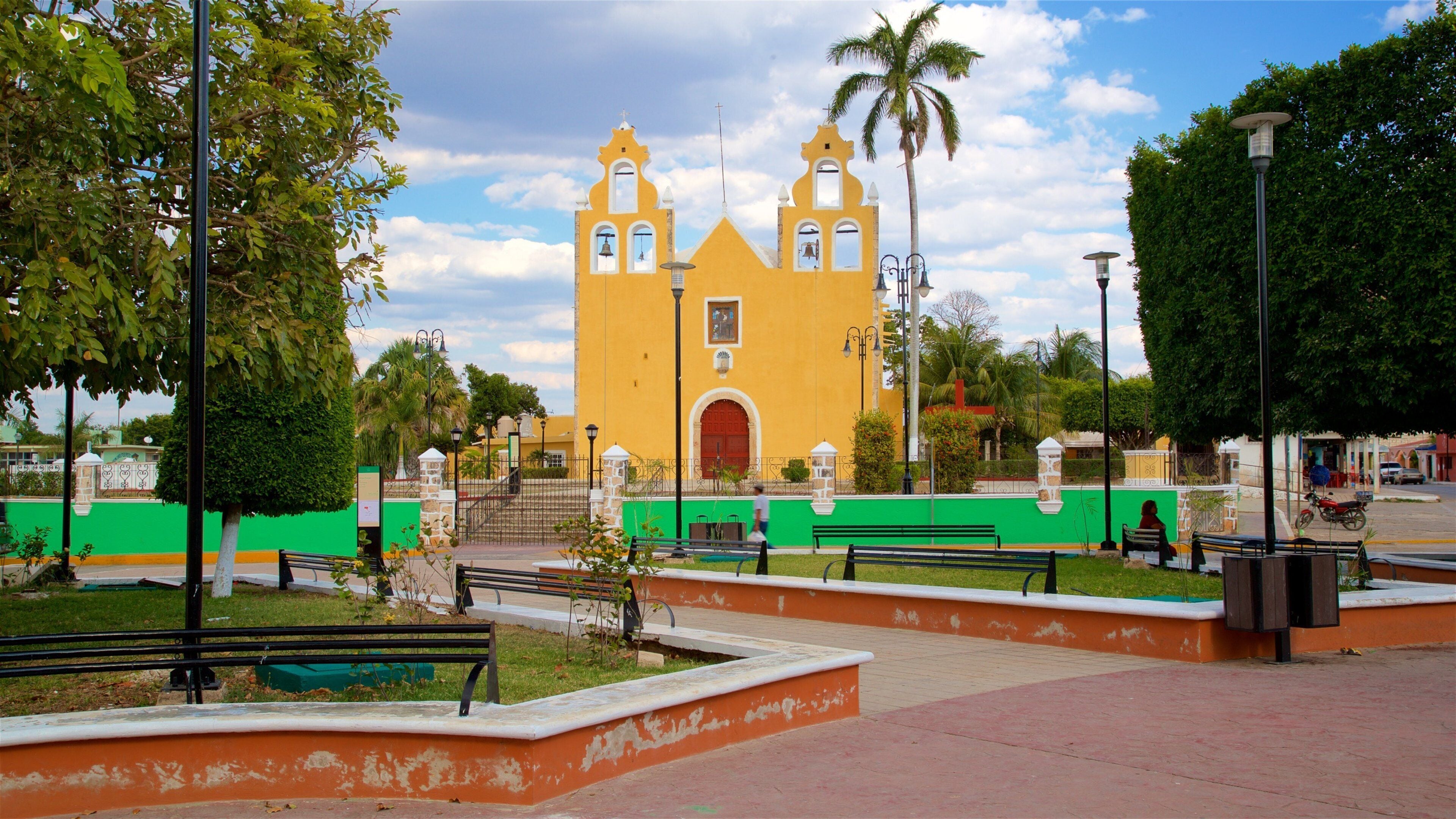 Igreja amarela com duas torres, rodeada por muros verdes e bancos.