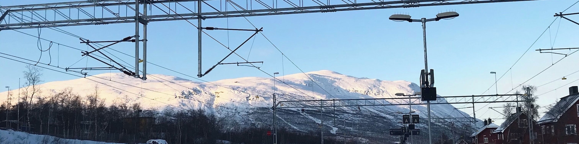 Trilhos de trem cobertos de neve, com linhas elétricas suspensas, uma montanha ao fundo e edifícios residenciais à direita.