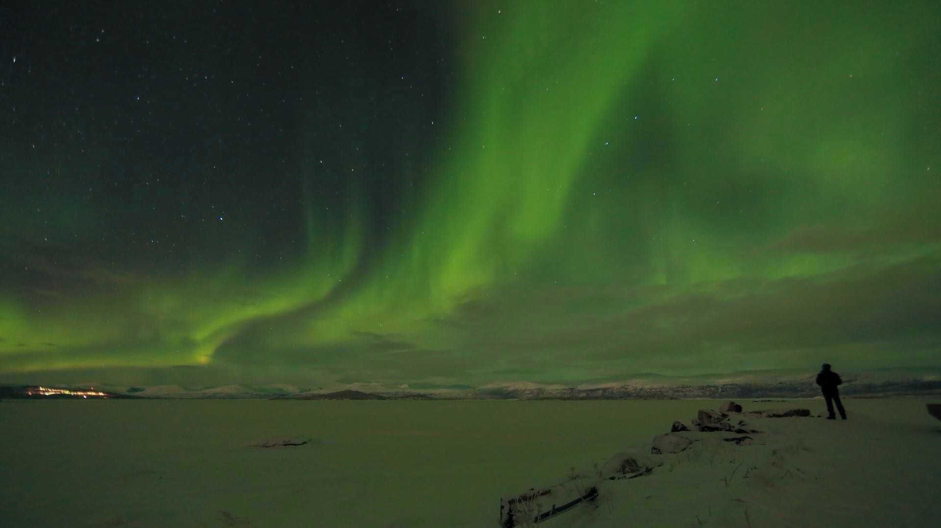Aurora boreal sobre uma paisagem nevada, com uma pessoa observando.