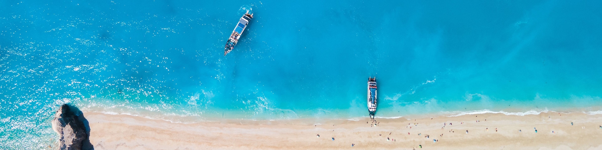 Uma praia com águas turquesa cristalinas, dois barcos e um afloramento rochoso.
