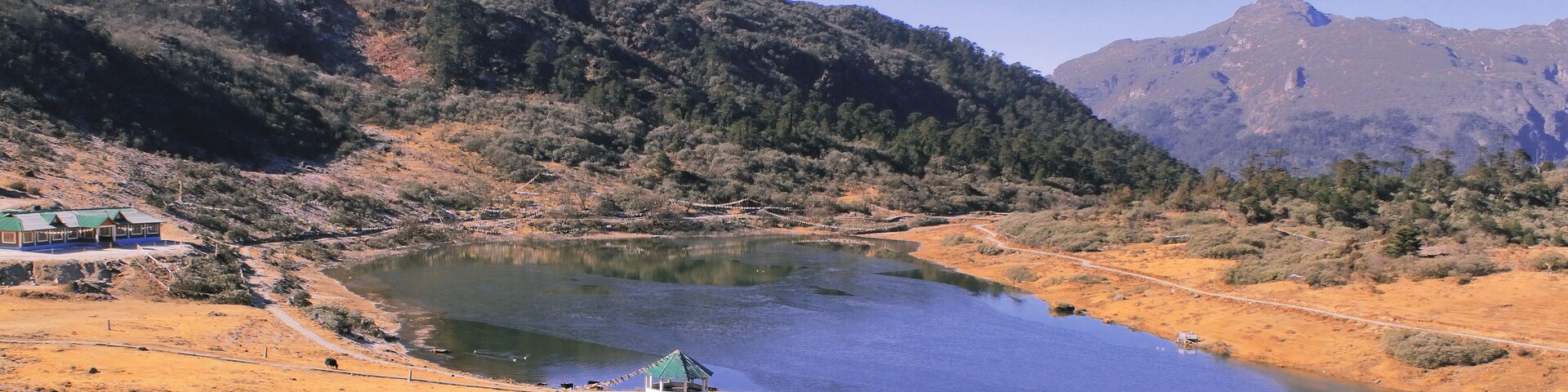 Um lago rodeado por montanhas, um pequeno edifício e um caminho de terra.