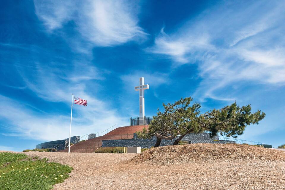 Uma cruz numa colina com uma bandeira, sob um céu azul.