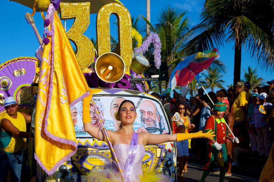Uma plataforma de desfile com um grande letreiro com o número '30', uma mulher num vestido roxo e uma multidão de pessoas.