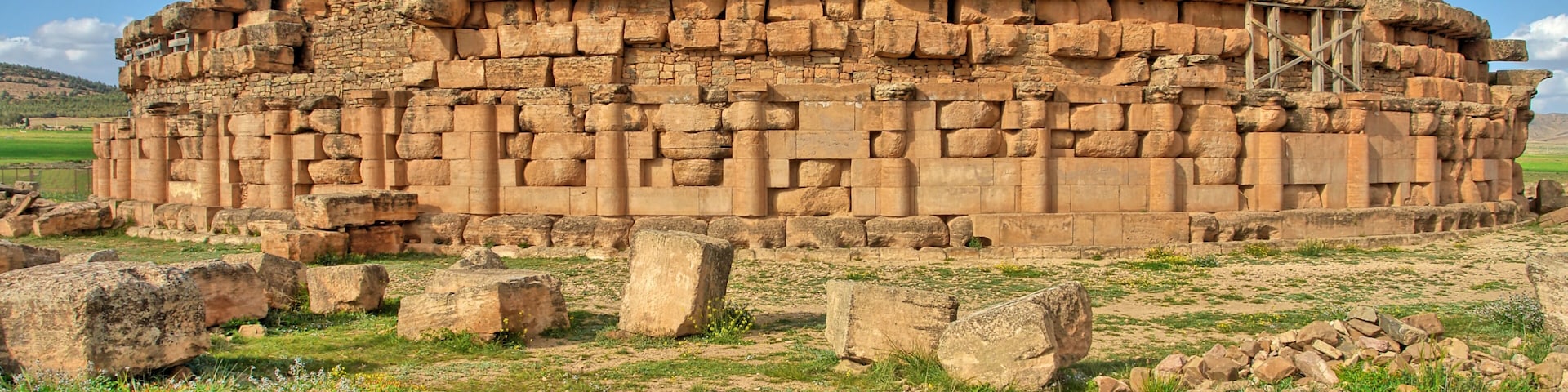 Uma grande estrutura de pedra antiga com topo arredondado, rodeada por um campo gramado e pedras espalhadas.
