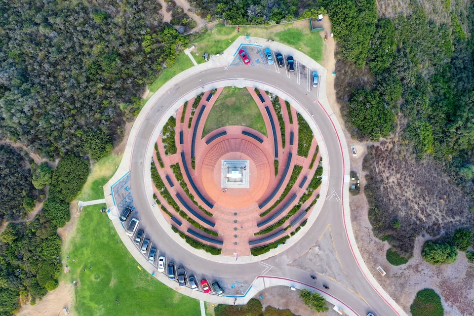Vista aérea de um rotatório com um monumento central, cercado por vegetação e carros estacionados.