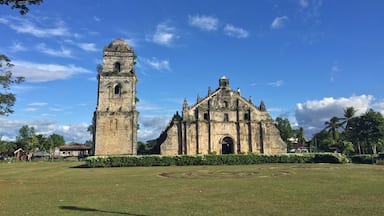 Uma igreja histórica com uma torre de sino e uma grande cúpula, rodeada por um jardim bem cuidado e palmeiras.