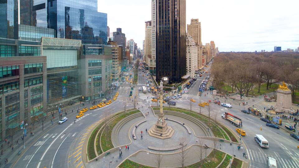 Um cruzamento urbano movimentado com um carrossel, edifícios altos e uma estátua no centro.