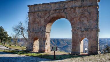 Um antigo arco de pedra com inscrições, rodeado por um espaço pavimentado e uma paisagem montanhosa.