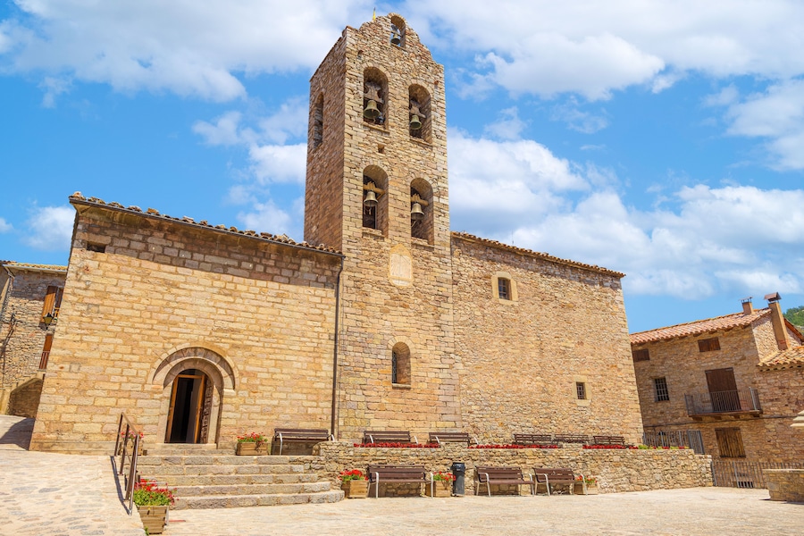 Um edifício histórico de pedra com uma torre de sino, degraus que levam à entrada e bancos com jardineiras floridas.