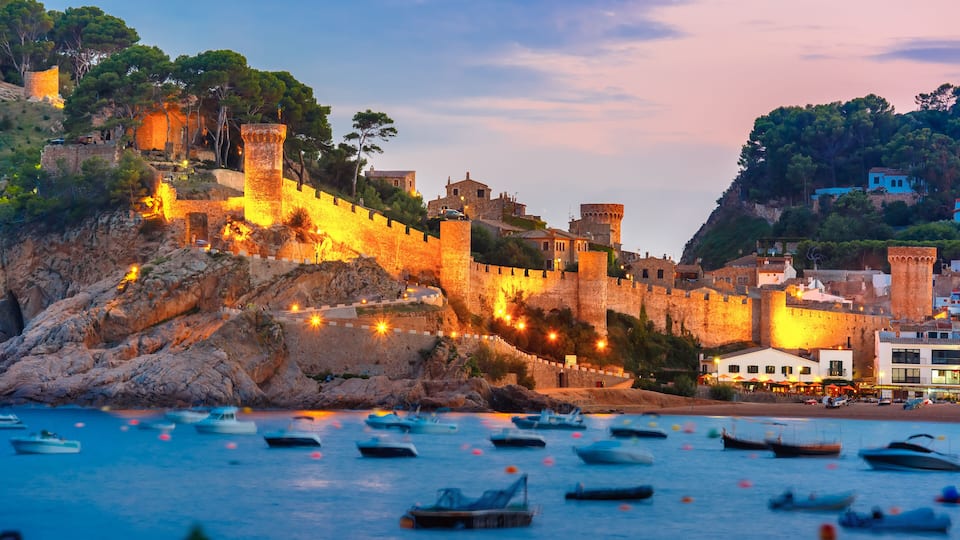 Uma cidade costeira com um castelo fortificado histórico, barcos na água e edifícios ao longo da colina.