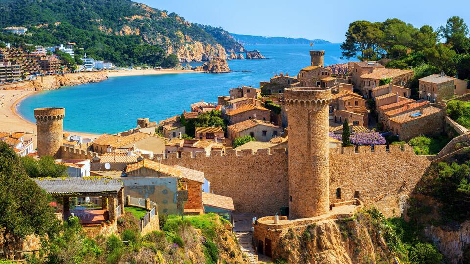 Uma vila costeira com um castelo imponente, uma praia e montanhas ao fundo.