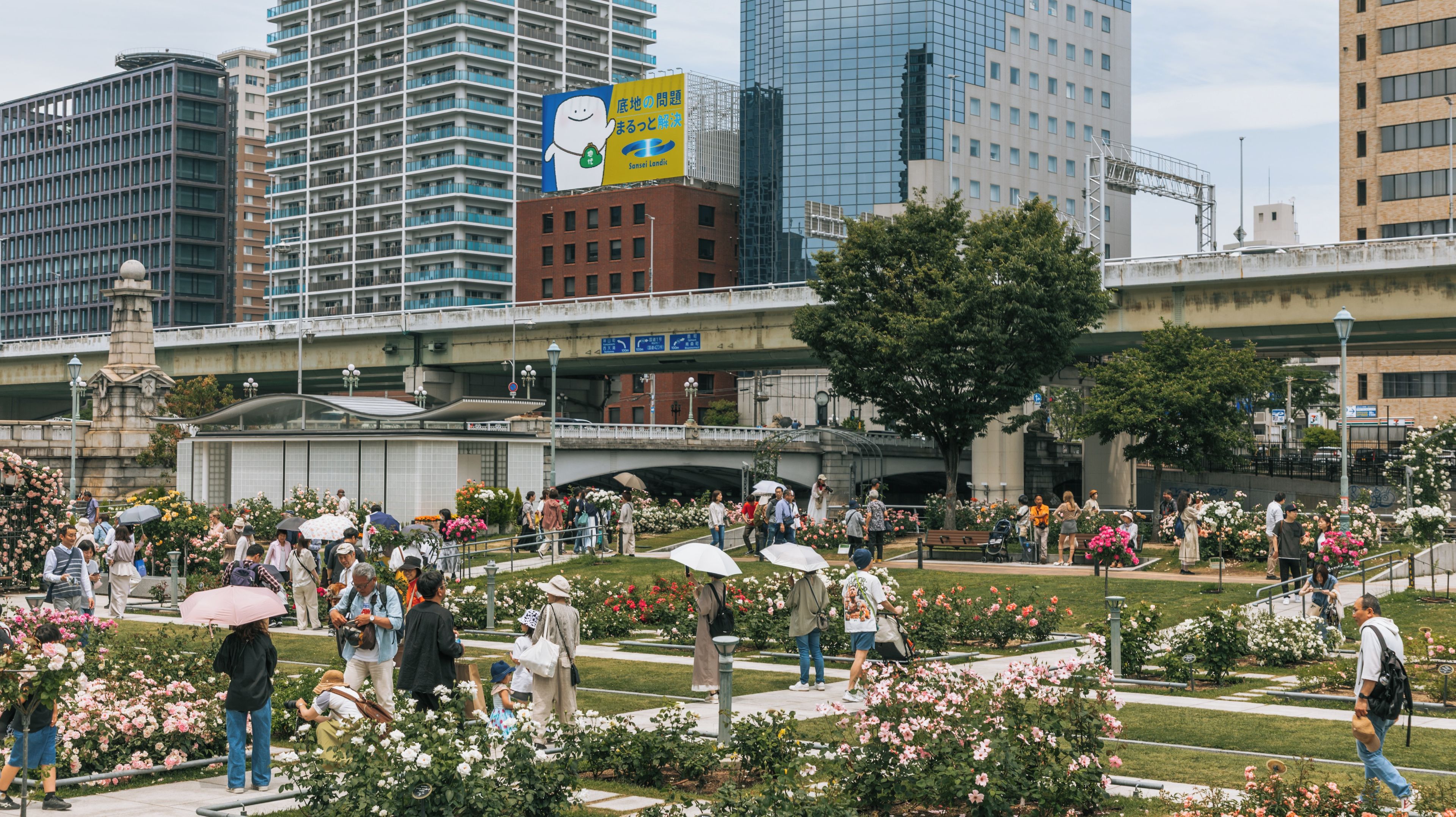 Um parque urbano movimentado com pessoas caminhando, algumas com guarda-chuvas, ao fundo edifícios modernos e um grande outdoor.