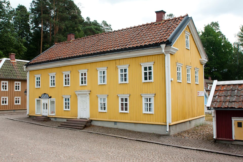 Um edifício amarelo com telhado de telhas vermelhas, janelas brancas e uma pequena construção vermelha nas proximidades.