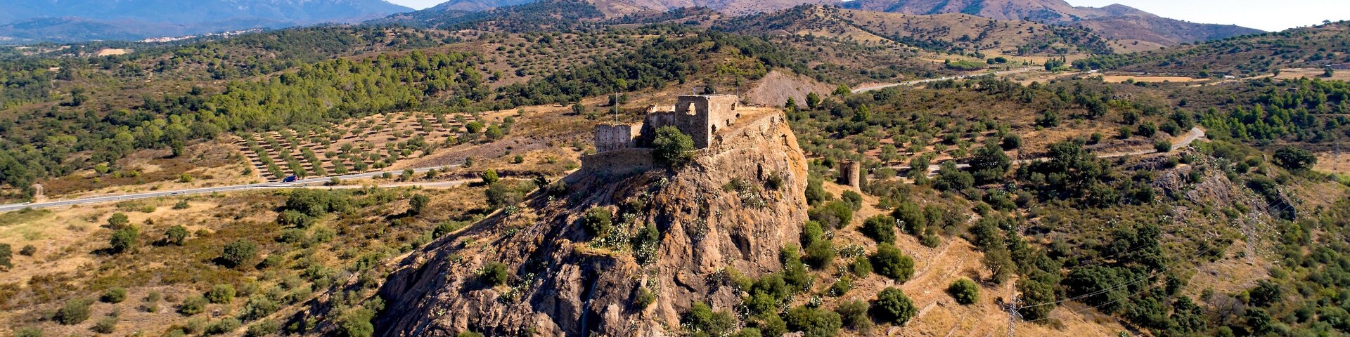 Uma fortaleza histórica numa colina, rodeada por montanhas e vegetação escassa.