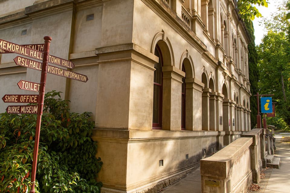 Um edifício histórico com um sinalizador indicando direções para Memorial Hall, RSL Hall, Town Hall, Centro de Informações para Visitantes, Sanitários, Escritório da Prefeitura e Burke Museum.