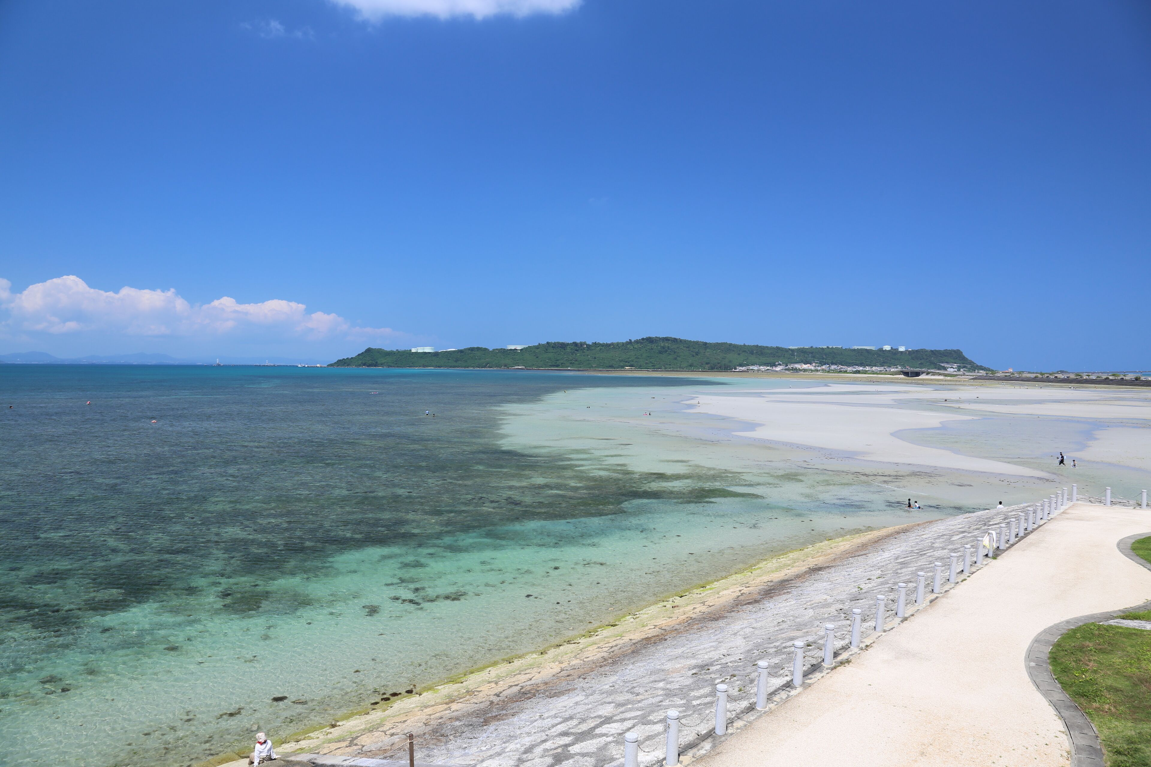Paisagem costeira com águas cristalinas, uma praia arenosa e uma colina verde ao longe.