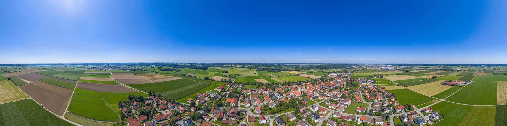 Vista aérea de uma cidade rural com campos verdes, casas de telhado vermelho e céu azul claro.