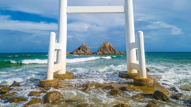 Um torii branco na praia, com ondas quebrando contra rochas e uma ilha rochosa ao fundo.