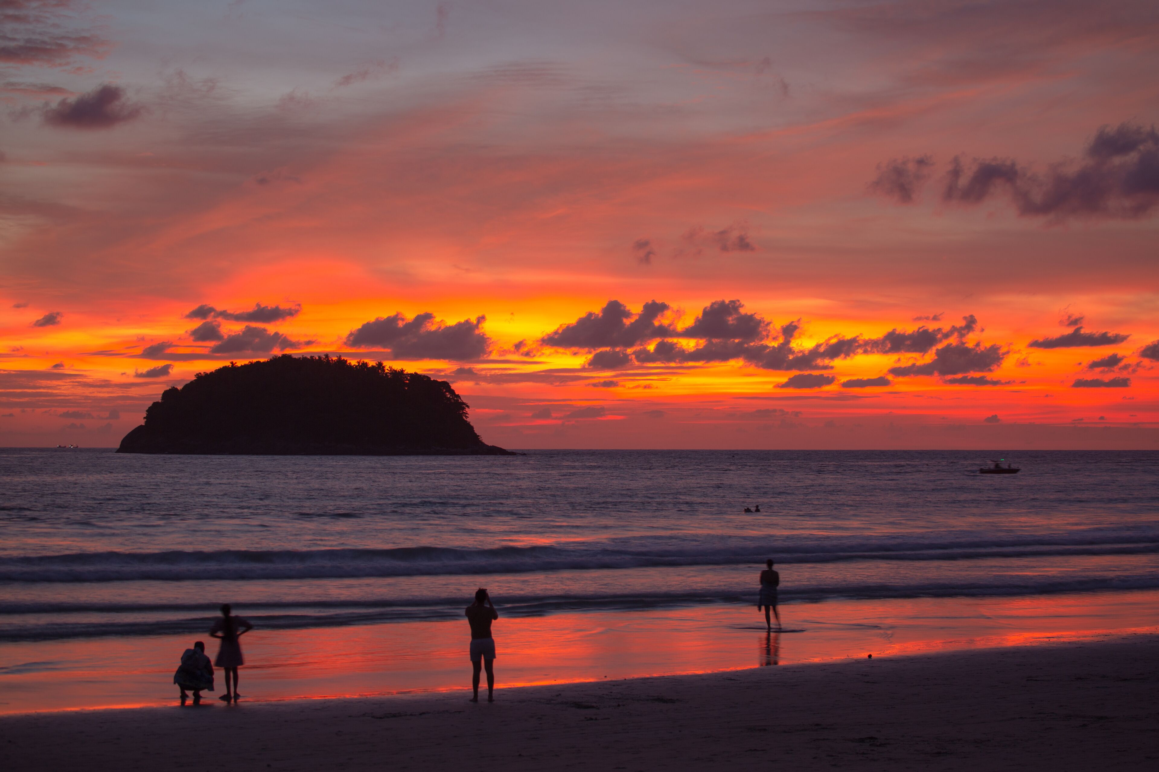 Um pôr do sol sobre uma praia, com silhuetas de pessoas e um barco na água.
