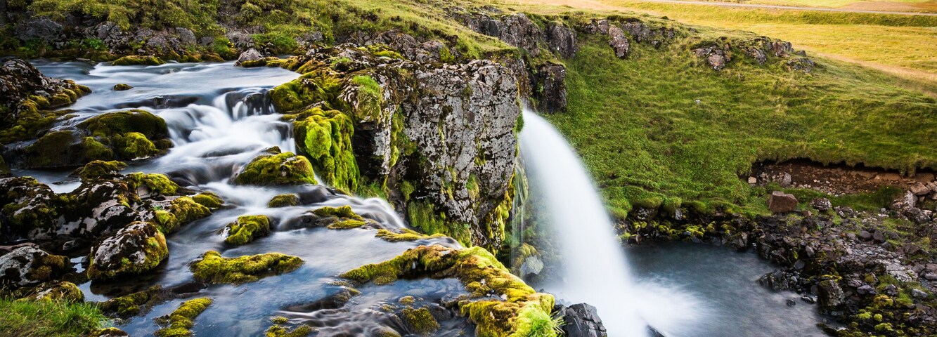Uma cascata descendo sobre rochas cobertas de musgo, um rio serpenteando por um vale verde, e uma montanha ao fundo.