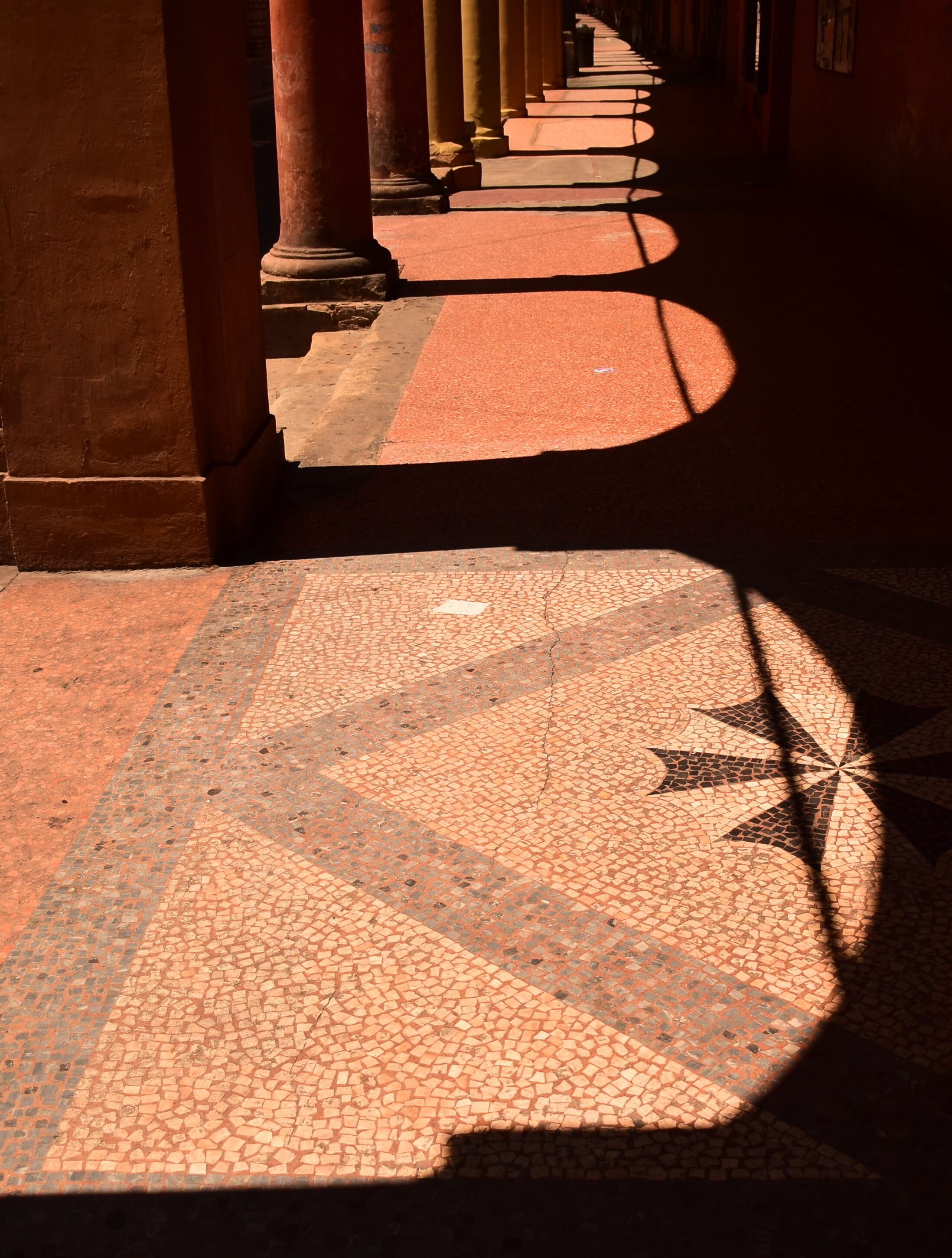 Um caminho pavimentado com azulejos formando um padrão de estrelas, colunas e um percurso banhado pelo sol.