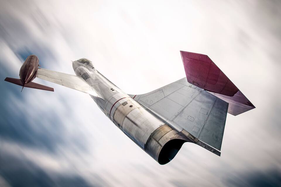 Uma aeronave de alta altitude com cauda vermelha e fuselagem branca voando por um céu nublado.