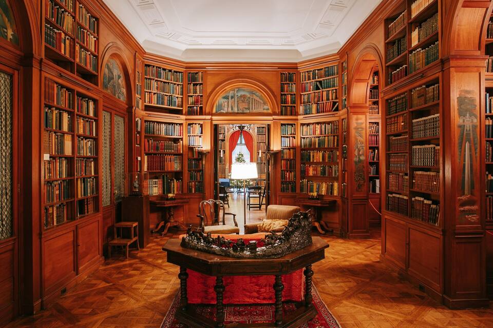 Uma biblioteca imponente com detalhes intrincados em madeira, estantes altas e uma área de leitura central.