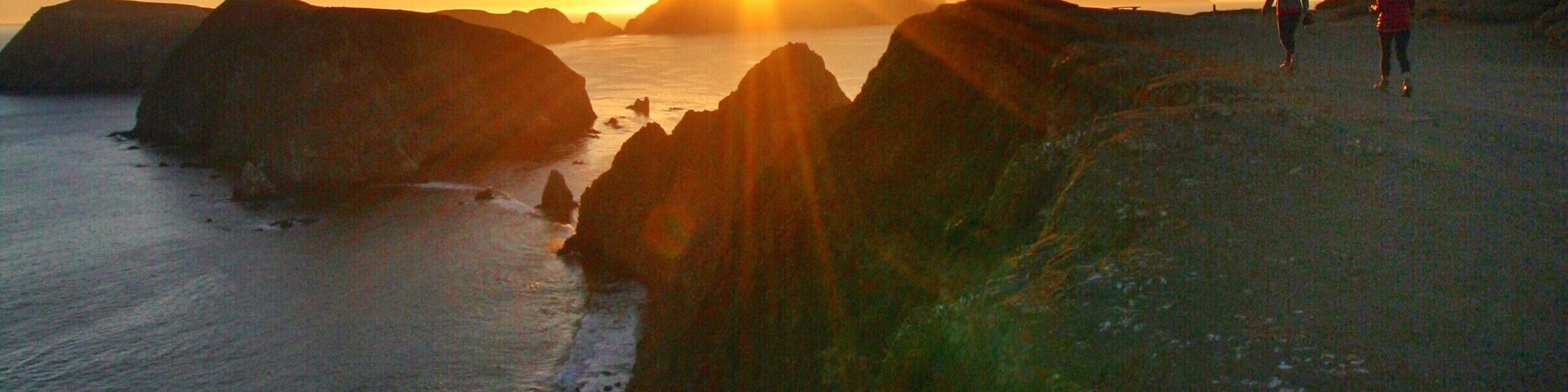 Um pôr do sol sobre uma costa rochosa com duas pessoas caminhando na praia.