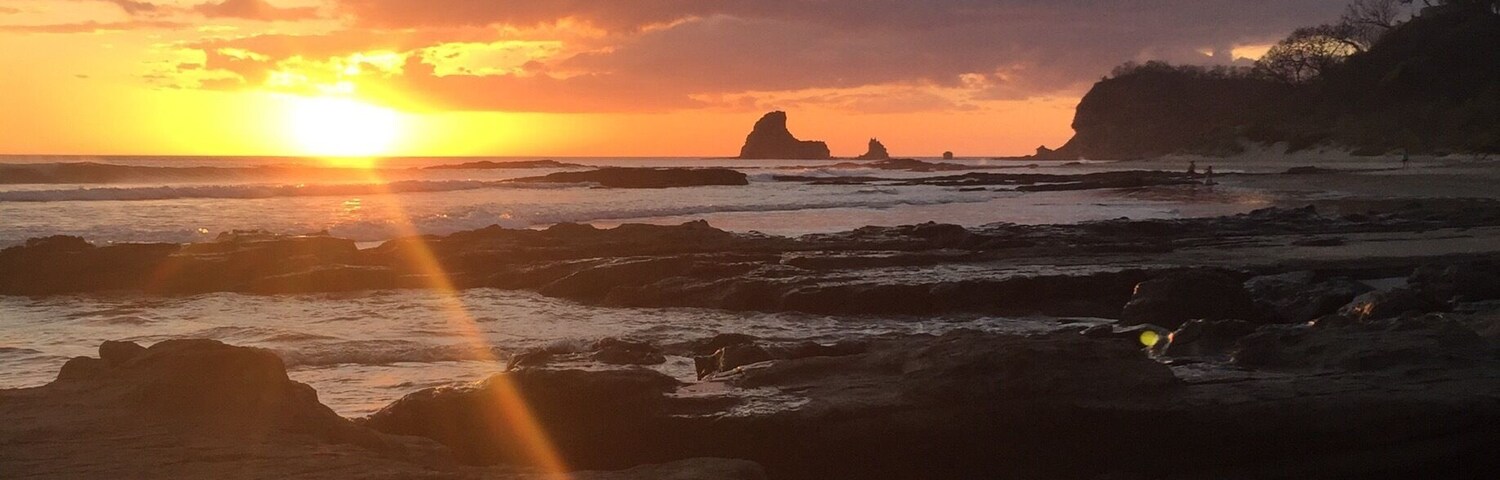 Um pôr do sol sobre um litoral rochoso, com ondas quebrando na praia e um céu limpo com algumas nuvens.