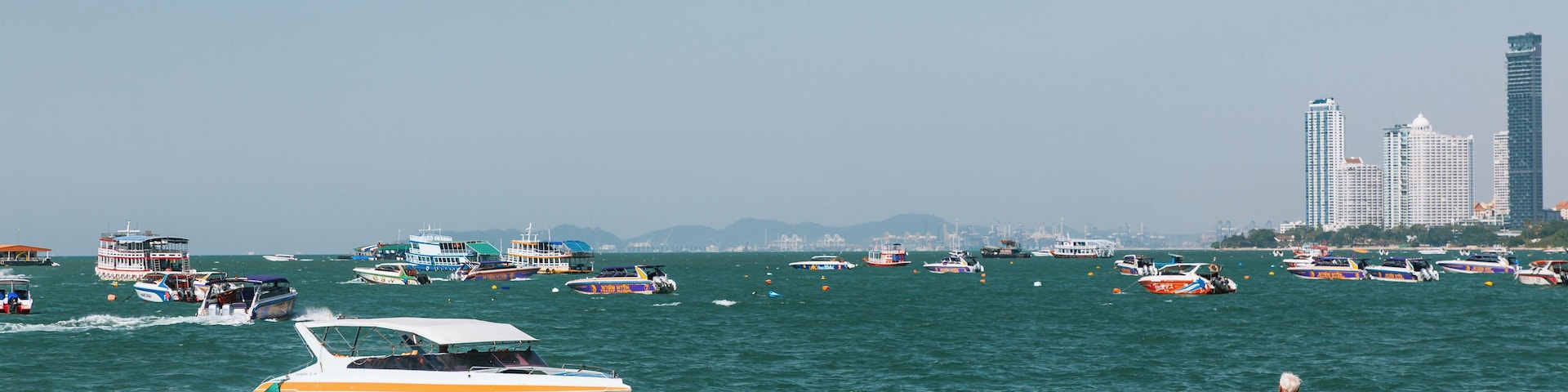 Um movimentado porto com vários barcos, uma praia de areia e um horizonte urbano ao fundo.