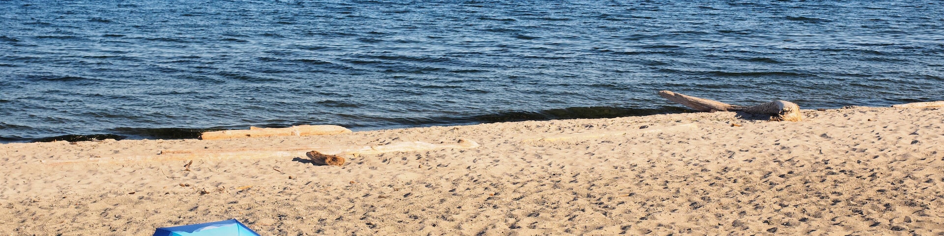 Uma praia arenosa com guarda-sol azul, mar calmo e montanhas ao longe.