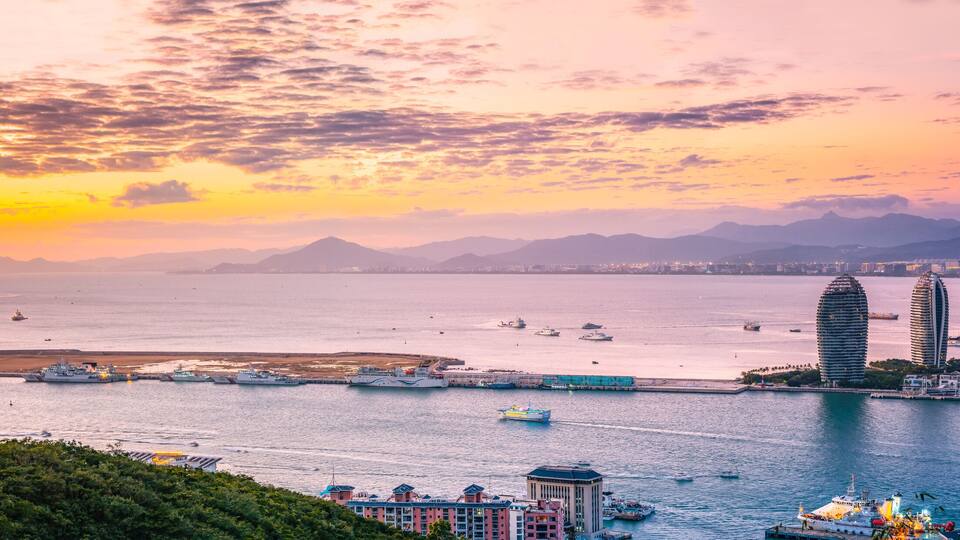 Paisagem costeira ao pôr do sol, com um porto importante, navios militares e imponentes arranha-céus modernos.