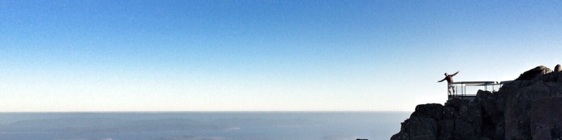 Paisagem rochosa com uma pessoa em um penhasco, observando um vasto vale e o horizonte distante.