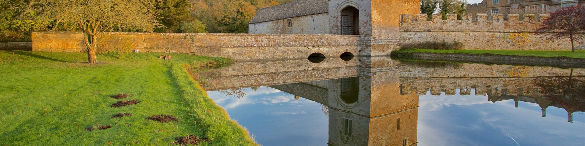Uma torre histórica com fossado, rodeada por vegetação exuberante e um reflexo límpido na água.