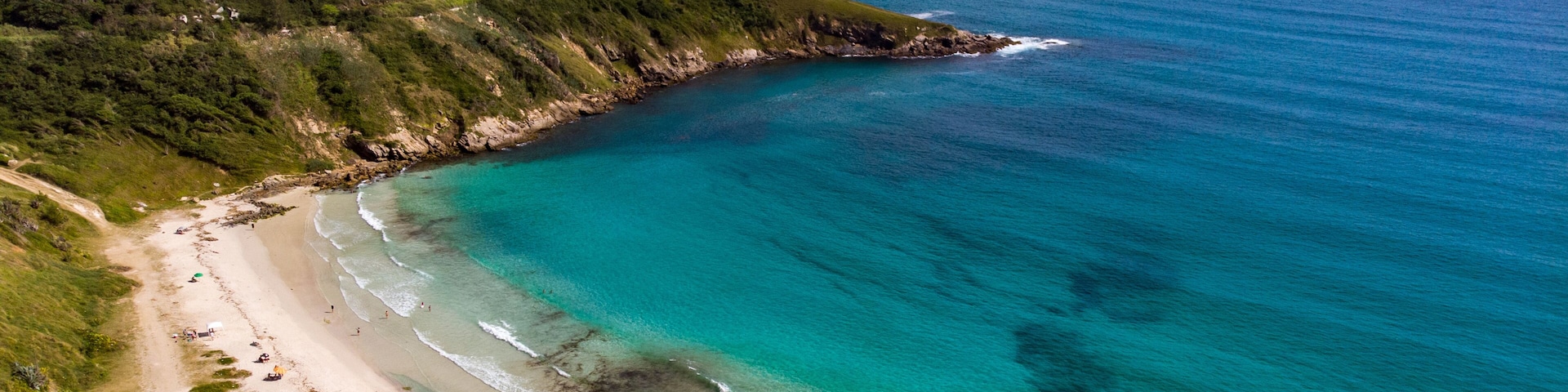 Paisagem costeira com praia, águas turquesa cristalinas e um relevo acidentado ao fundo.