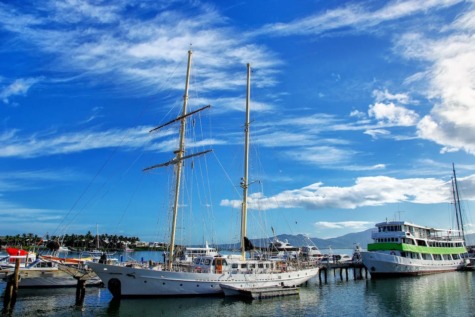 Uma marina com vários veleiros atracados, um ferry verde e branco e uma paisagem montanhosa ao fundo.