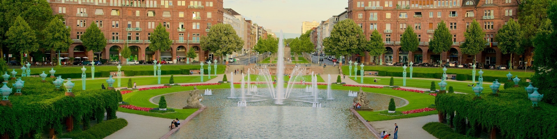 Uma imponente fonte no centro de uma praça bem cuidada, rodeada por edifícios simétricos e vegetação exuberante.
