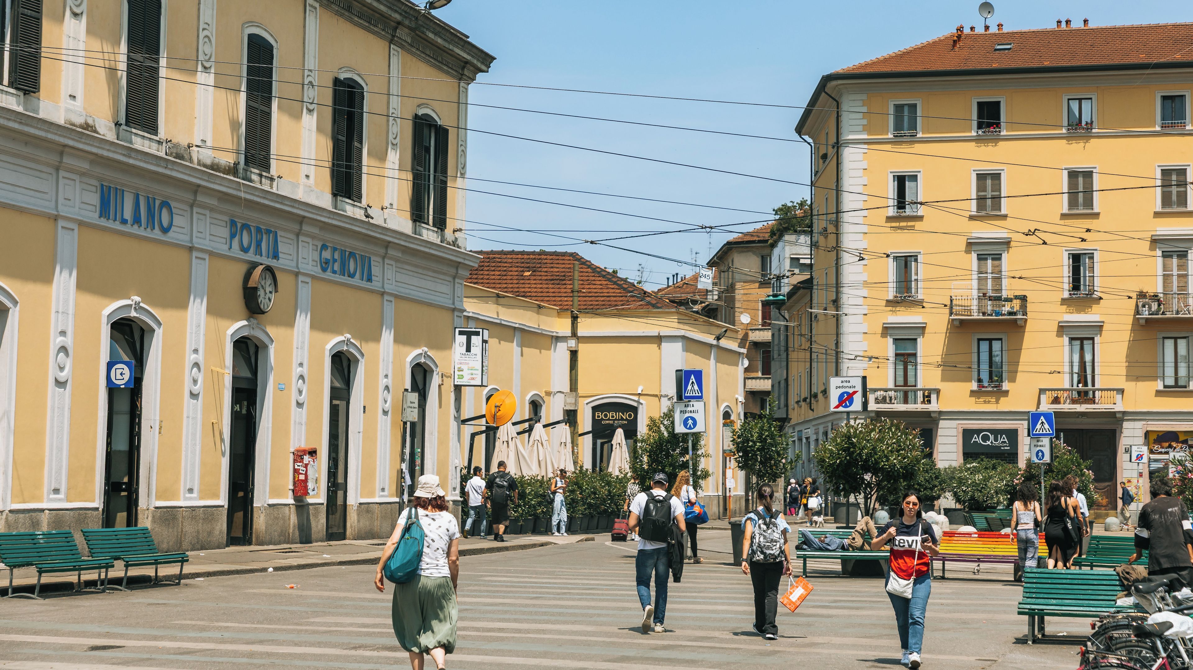 Rua com pedestres, bancos e edifícios com placas como 'MILANO PORTA GENOVA' e 'AQUA'.