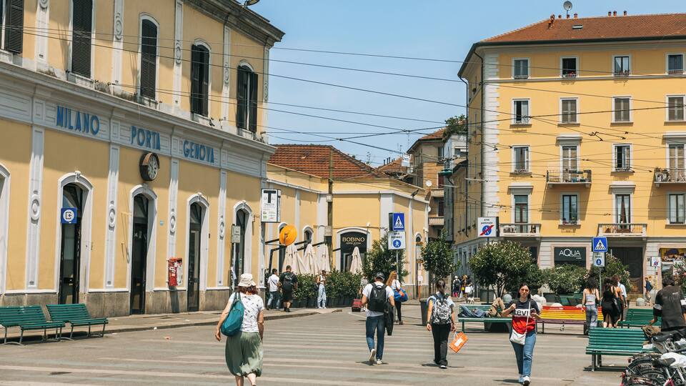 Rua com pedestres, bancos e edifícios com placas como 'MILANO PORTA GENOVA' e 'AQUA'.