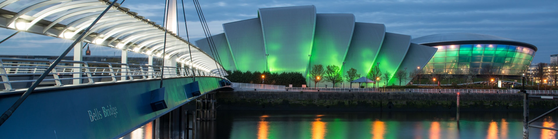 Uma ponte com a inscrição 'Bells Bridge', um edifício moderno com iluminação verde e uma massa de água calma refletindo as estruturas.